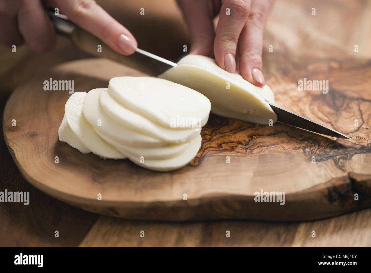 Female Teen Hand Slicing Mozzarella Cheese With Knife On Wooden Board
