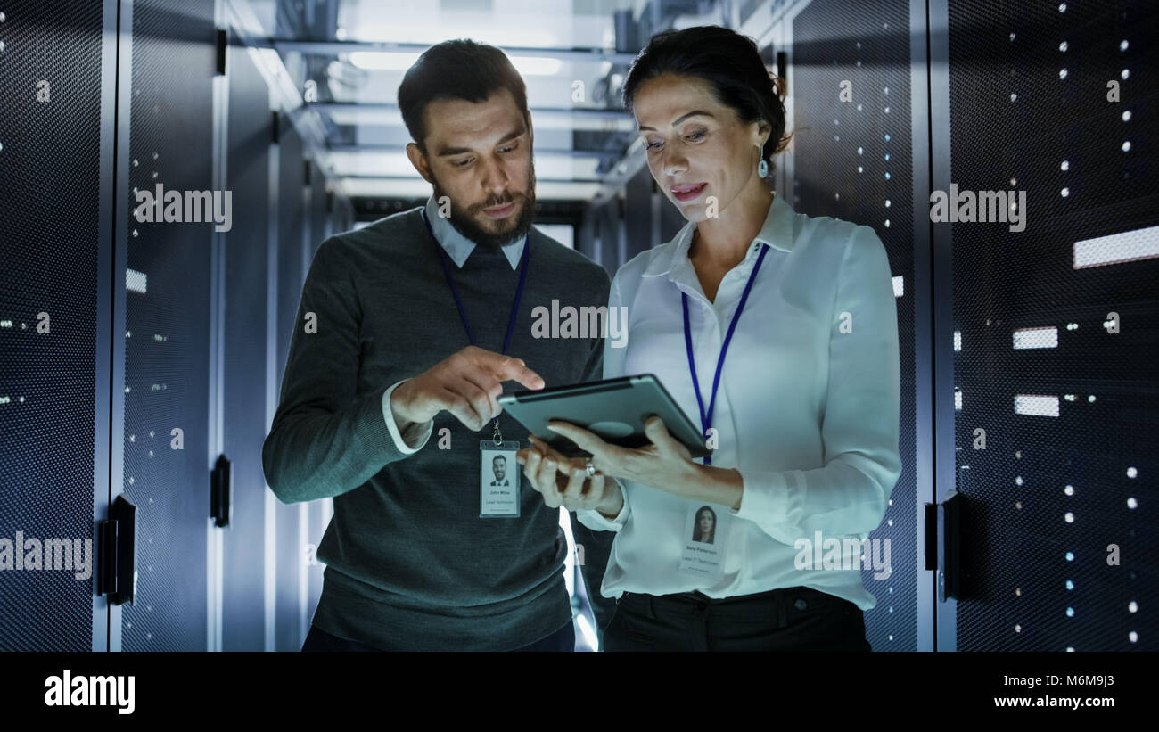Two IT Engineers Walking in Data Center / Server Room, Talking, Running Diagnostics with Tablet ...