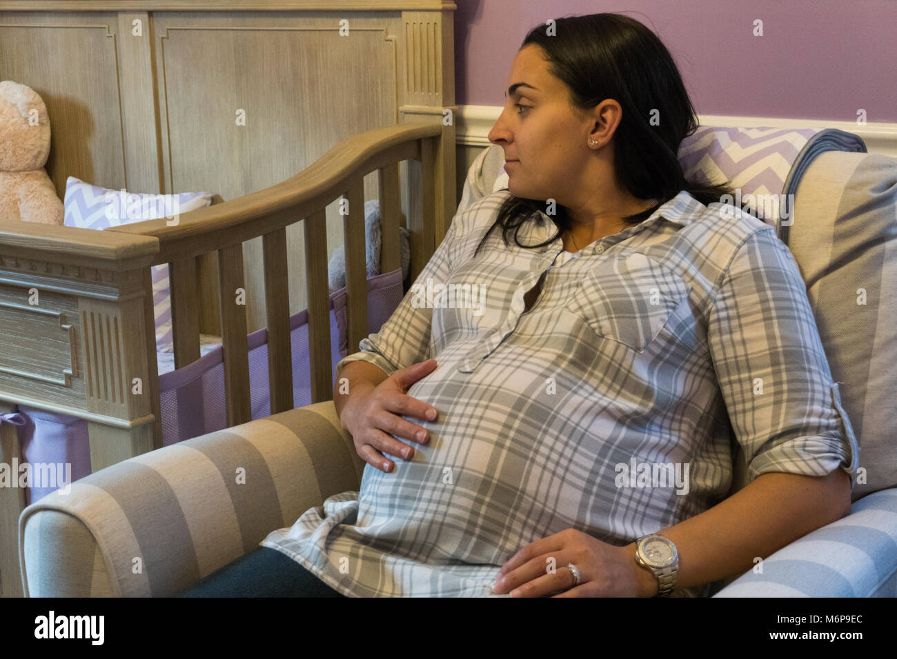 Beautiful pregnant woman sit in rocking chair next to crib touching ...
