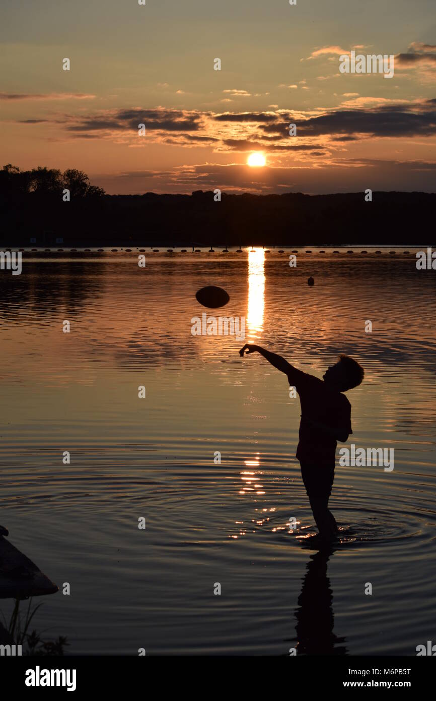 Boy throwing rugby ball in a lake with sun setting giving reflections ...