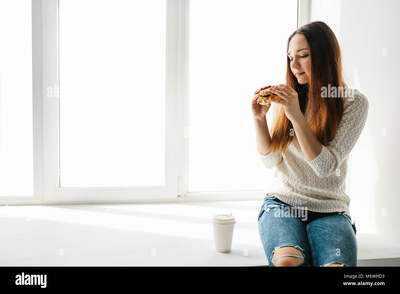 A young beautiful girl sits on a window and eats a burger with coffee ...