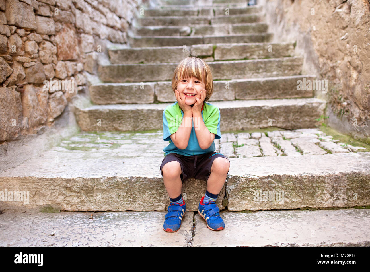 cute smiling blonde little boy sitting on old cobblestone stairs with ...