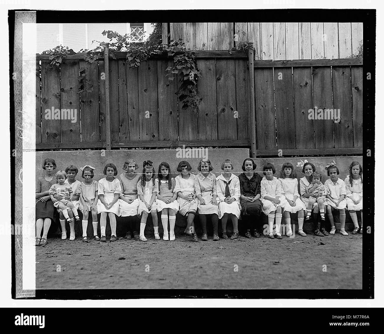 A photograph of children seated in front of a fence, captured on August ...