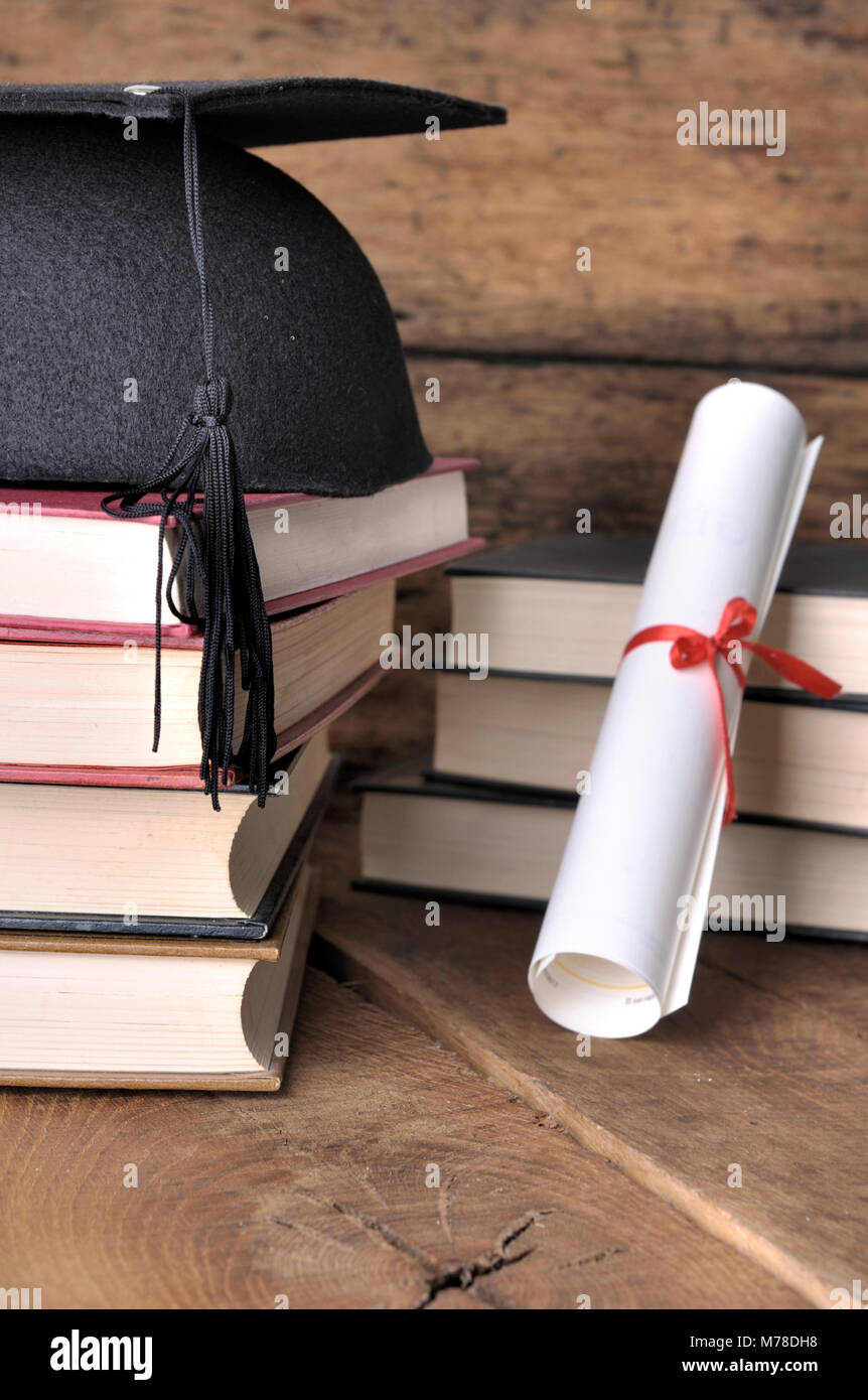 Graduate rolled up in a red ribbon and a black hat on pile of book on ...