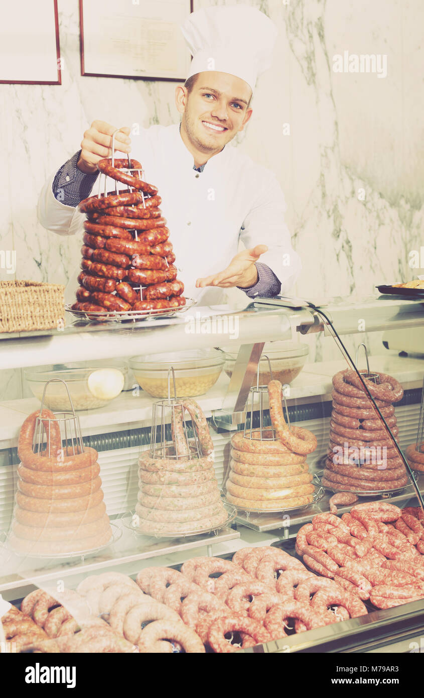 Glad young cook in white uniform at butcher store offering meat and ...