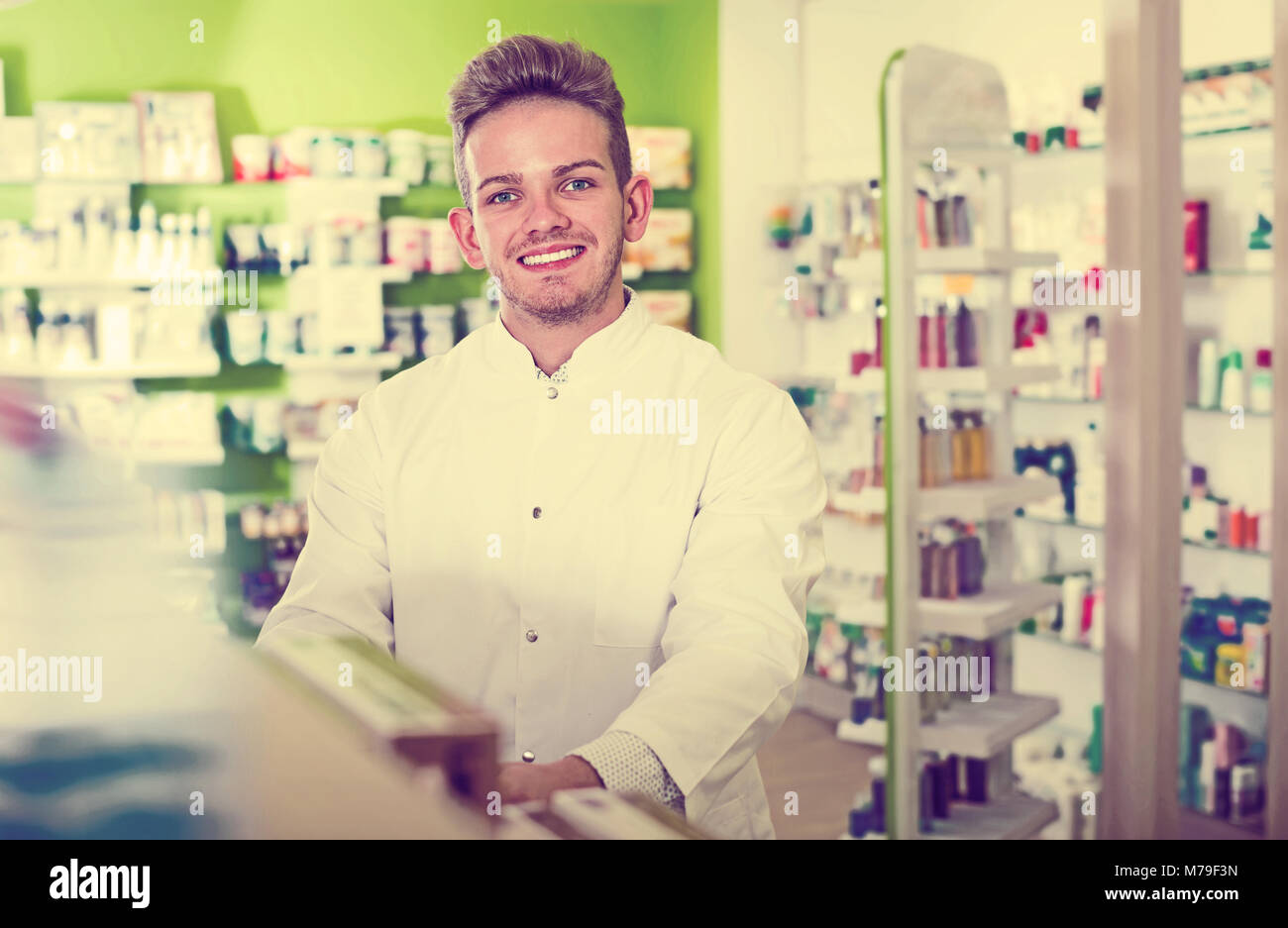 Positive pharmacist wearing white coat standing among shelves in drug ...