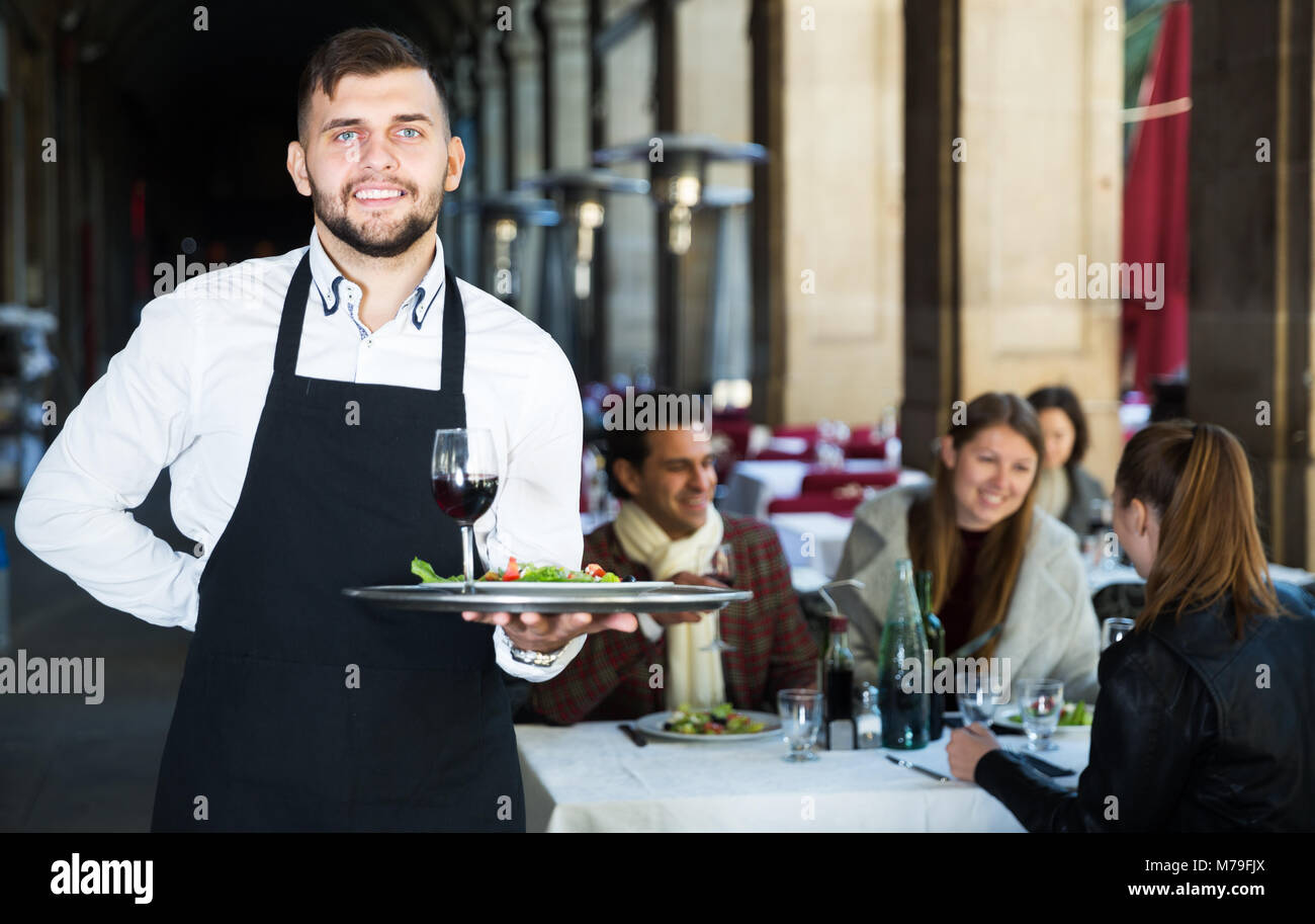 Polite waiter holding tray at restaurant with customers his behind ...