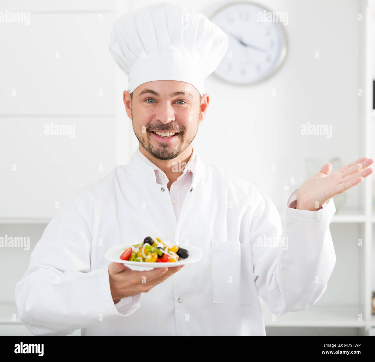 Young happy european cook in cap offering greek salad for lunch Stock ...