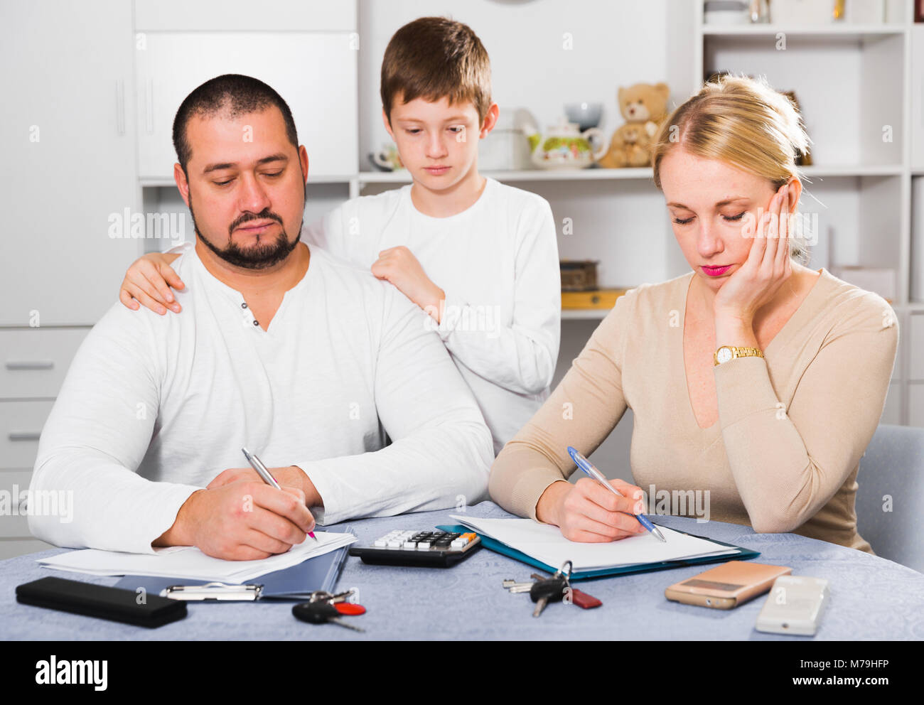 Tense parents writing documents at home table with sad son behind Stock ...