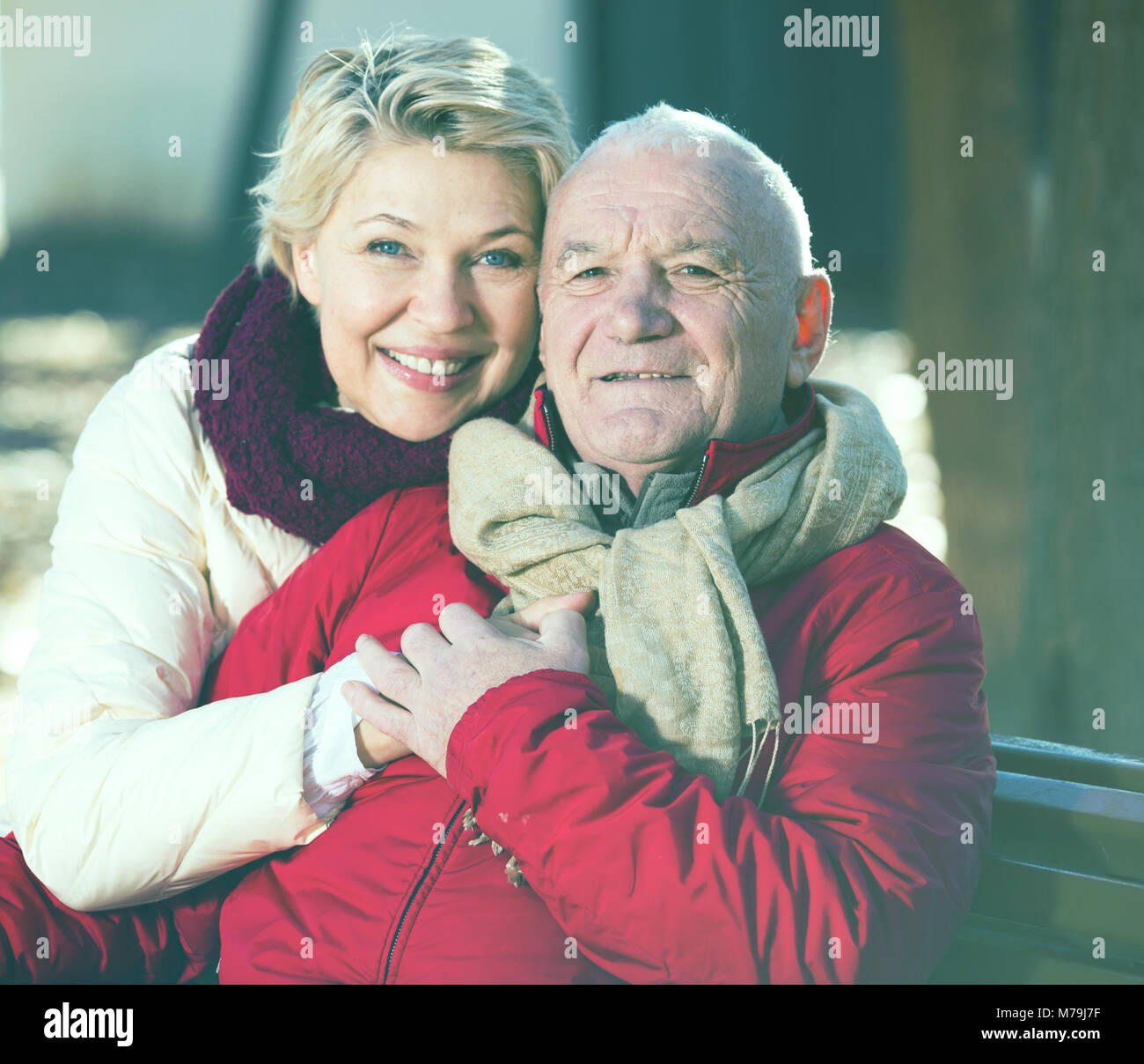 Aged husband and wife sitting together on bench in park on chilly day ...