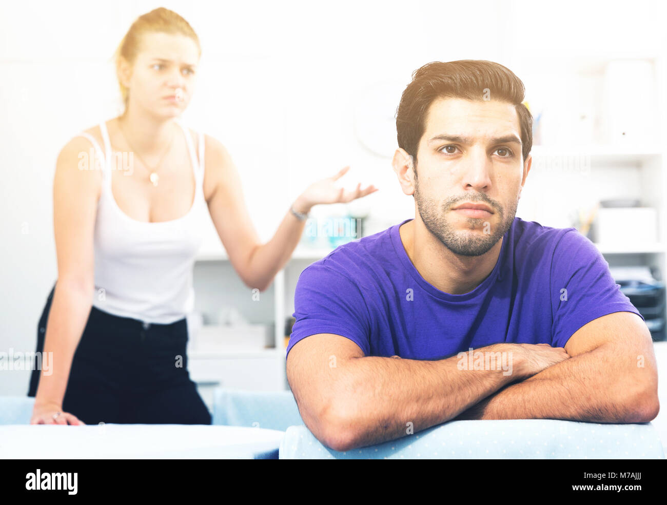 Upset young man sitting at home table with disgruntled woman behind ...