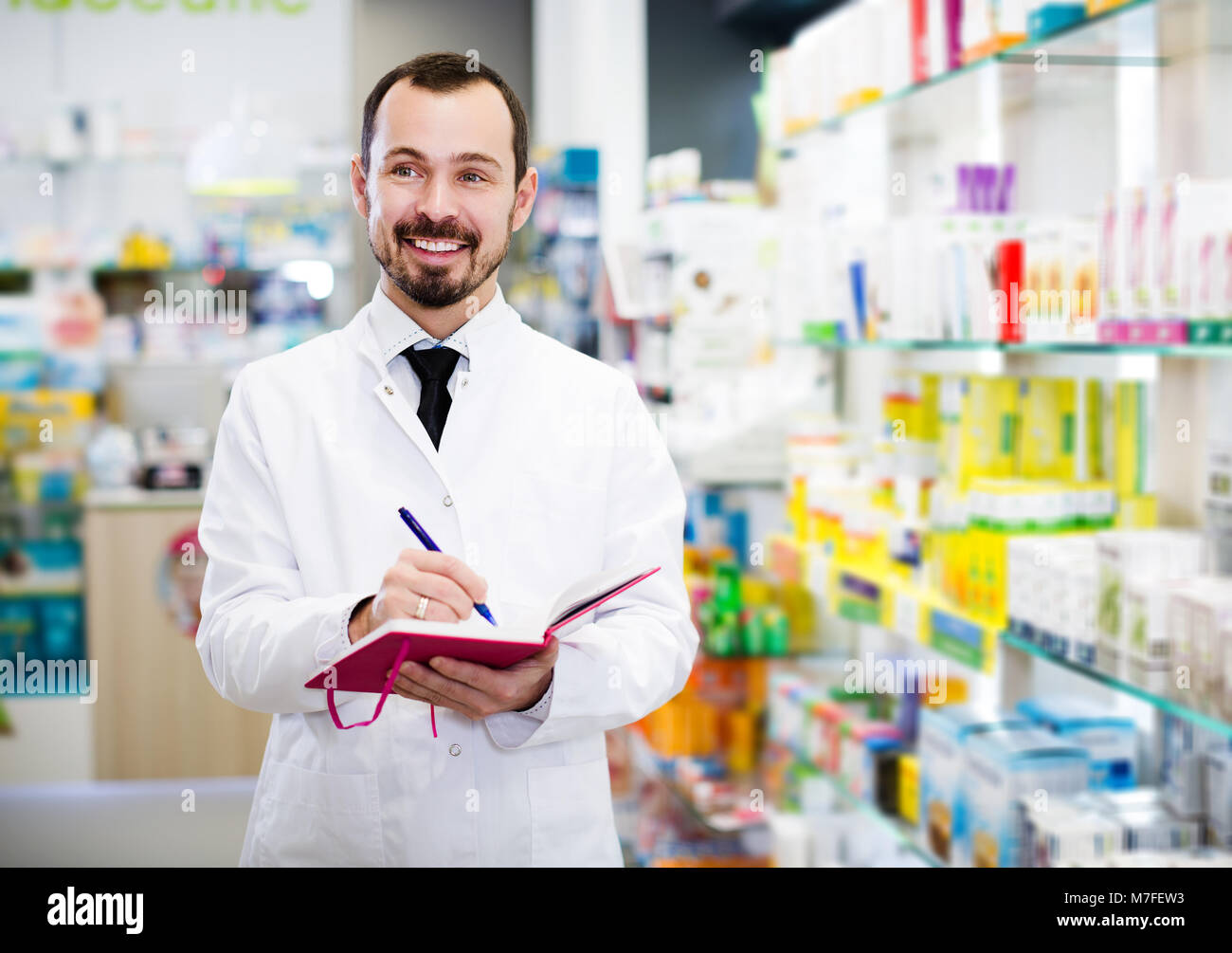 Smiling adult pharmacist writing down assortment of drugs in pharmacy ...