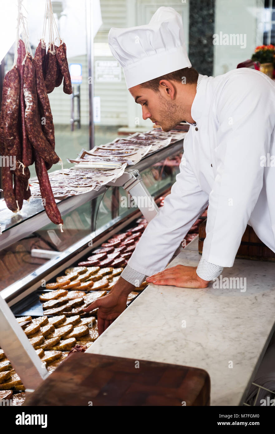 Smiling young cook in white uniform in grocery shop offering meat and ...