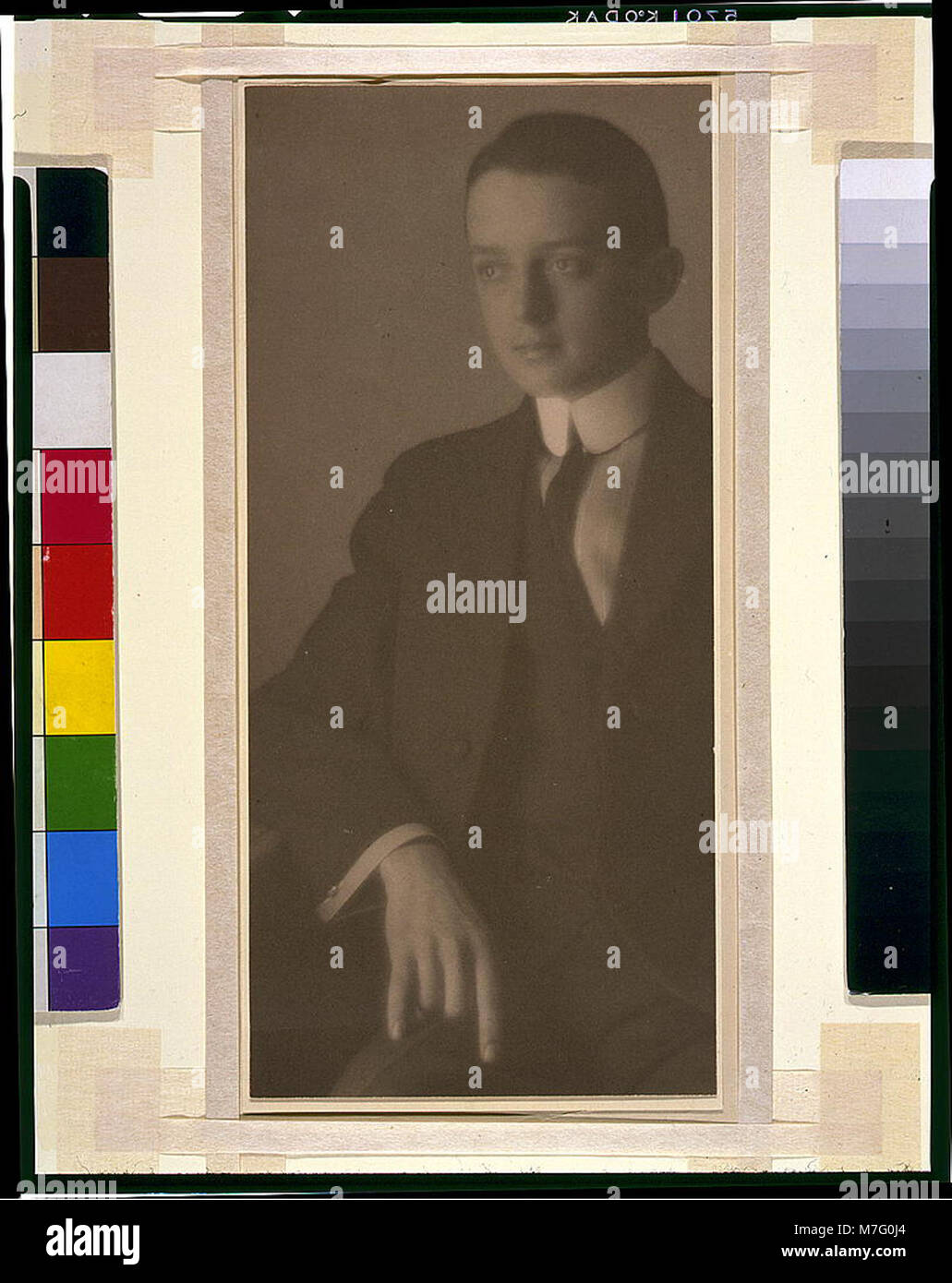 This photograph depicts a young man seated in a suit, with his hand ...