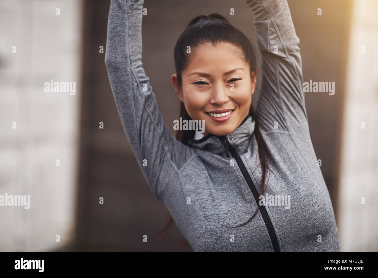 Fit young Asian woman in sportswear smiling while standing outside stretching her arms before ...