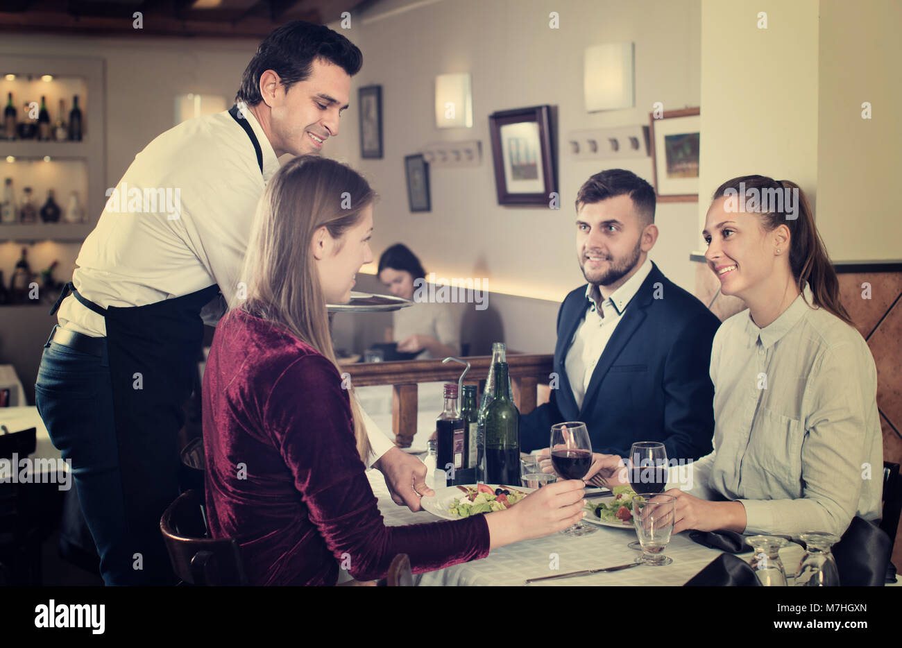 Positive waiter serving meals to friendly company guests at restaurant ...