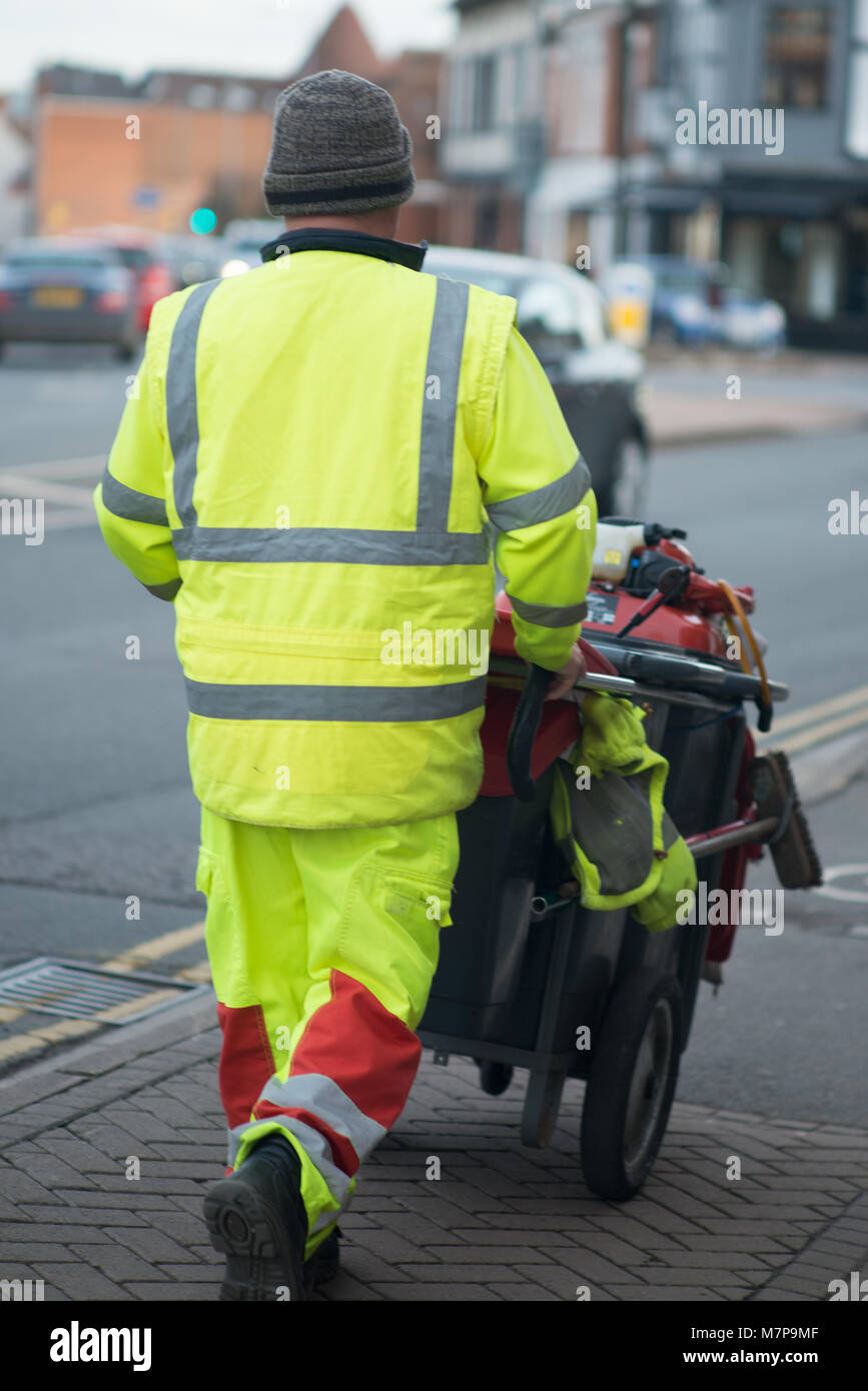 street sweeper walks down road pushing ref and grey barrow with ...