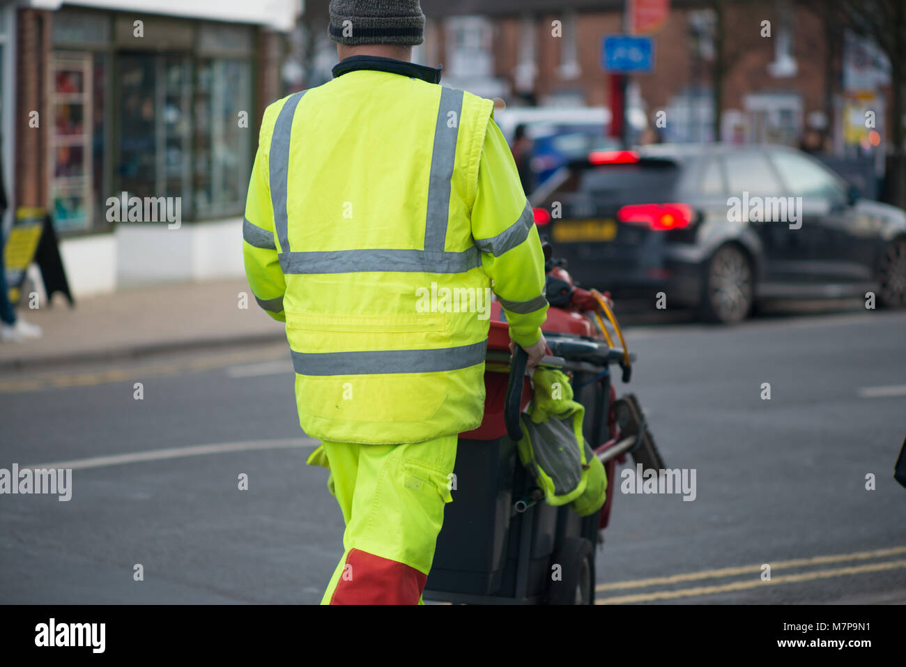 street sweeper walks down road pushing ref and grey barrow with ...
