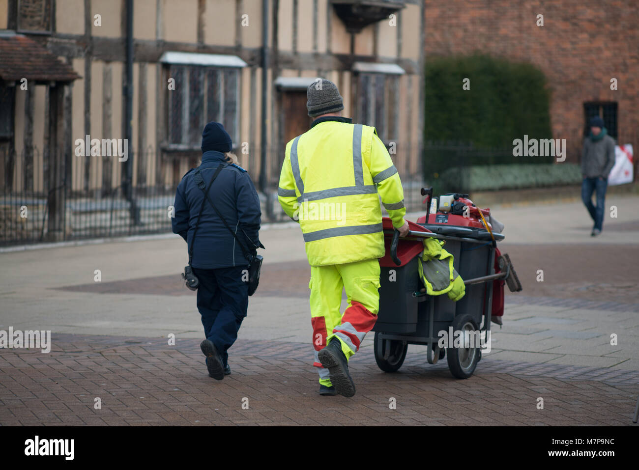 street sweeper walks down road pushing ref and grey barrow with ...