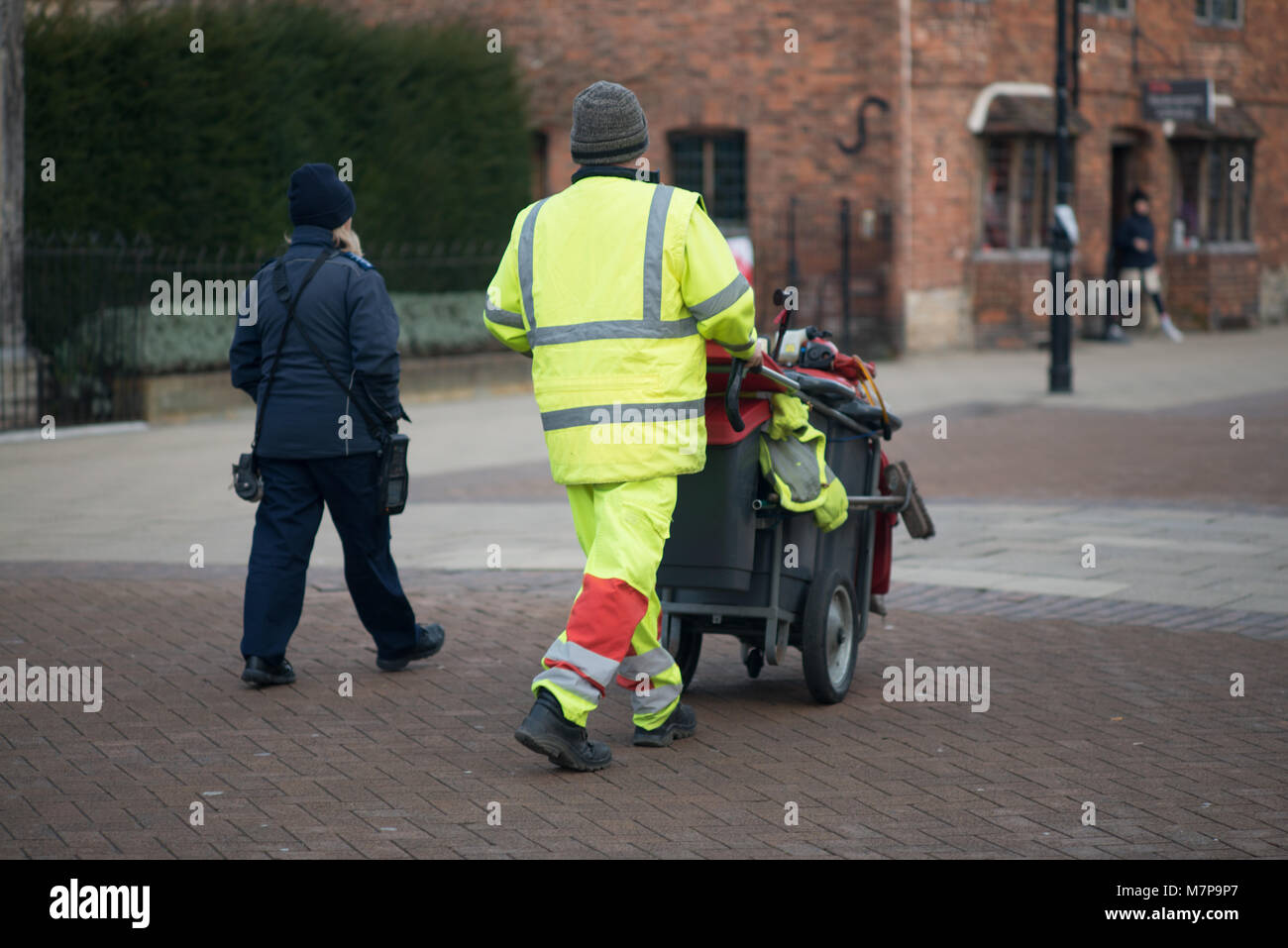 street sweeper walks down road pushing ref and grey barrow with ...