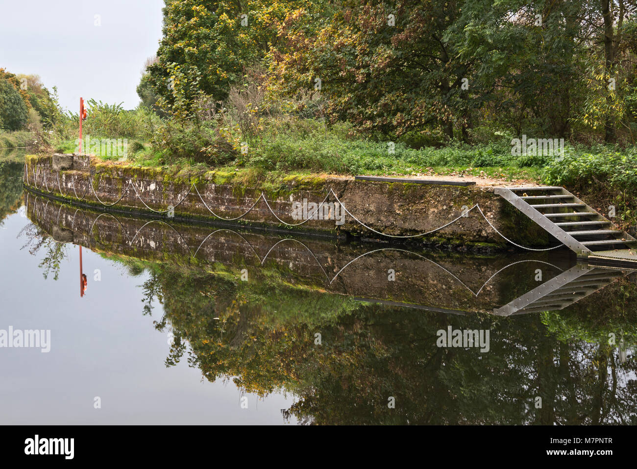 reflections of a stone quayside and trees in the still waters of the ...