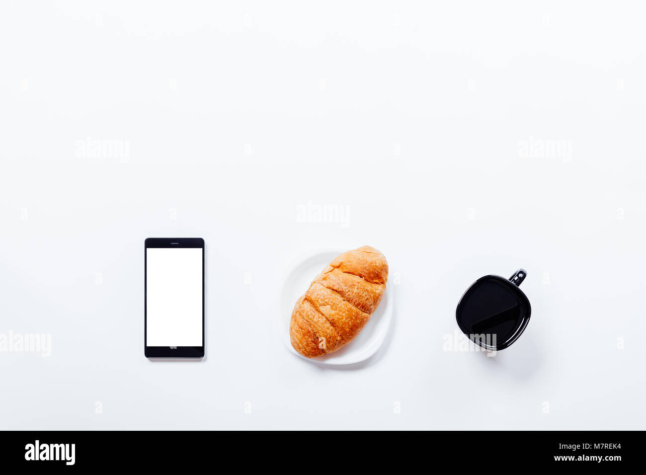 Mobile phone with blank screen, croissant and coffee on white table top ...