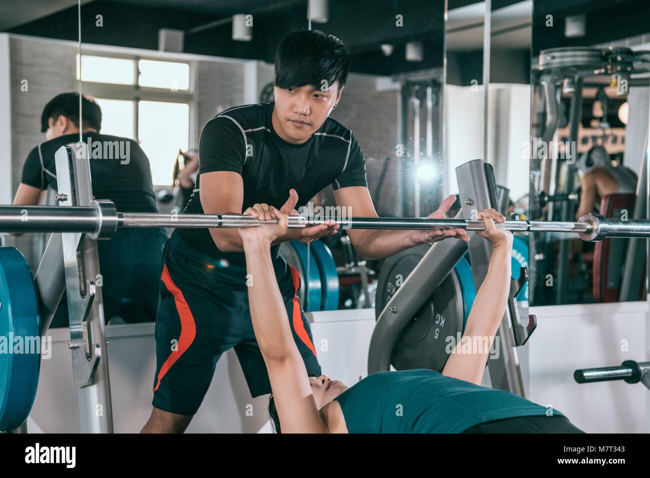 Asian male personal trainer helping a young woman lift a barbell while ...