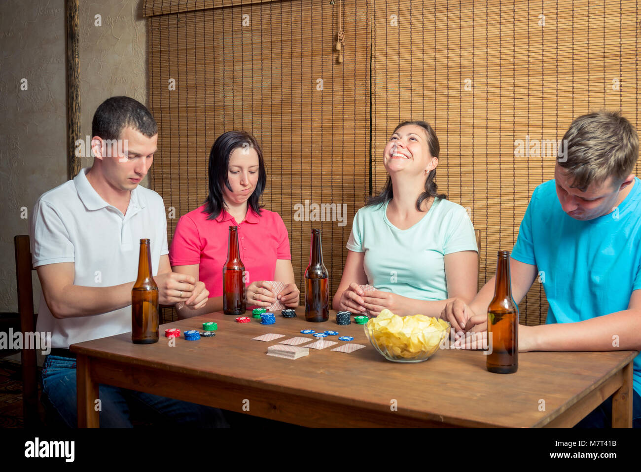 young friends playing cards sitting at a table indoors, drinking beer ...