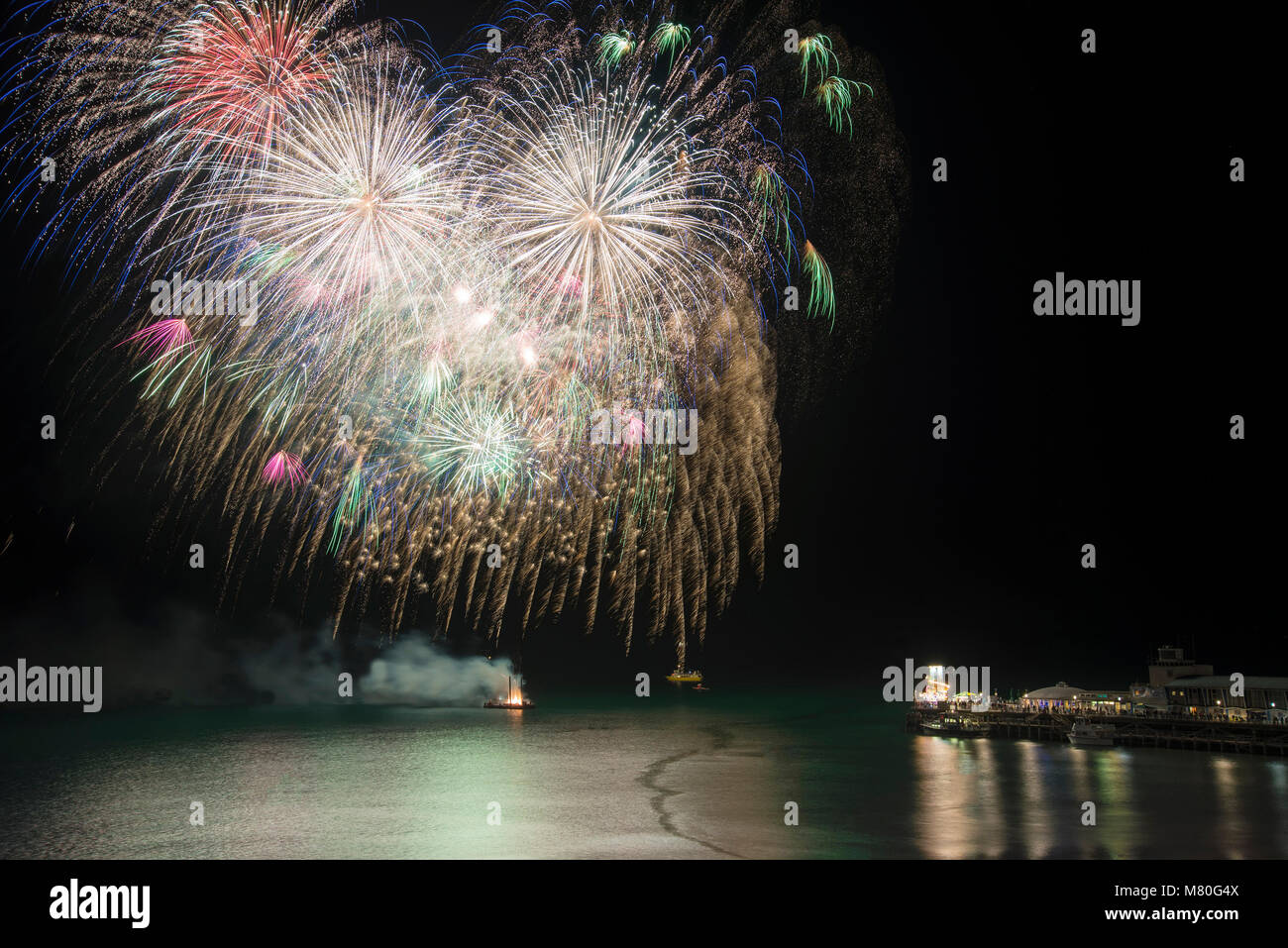 Fireworks display over sea with pier and boats in water landscape scene ...