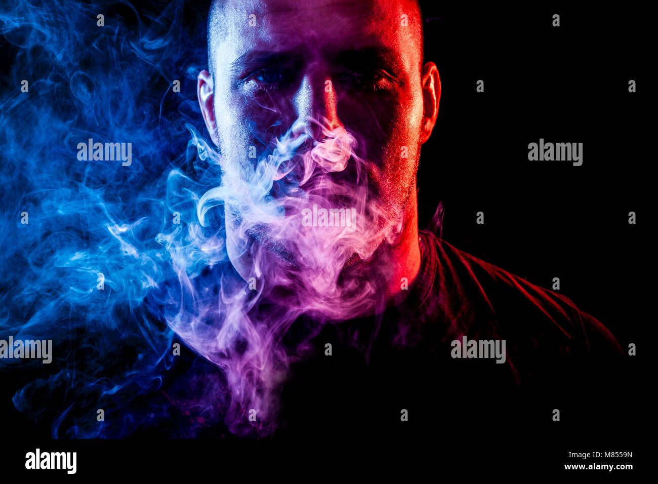 A young man exhales a cloud of colored smoke of blue and red color on a ...