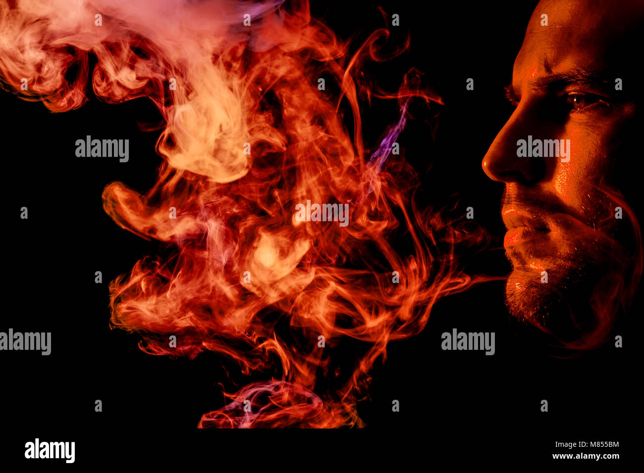 A young man exhales a cloud of colored smoke of red color on a black ...