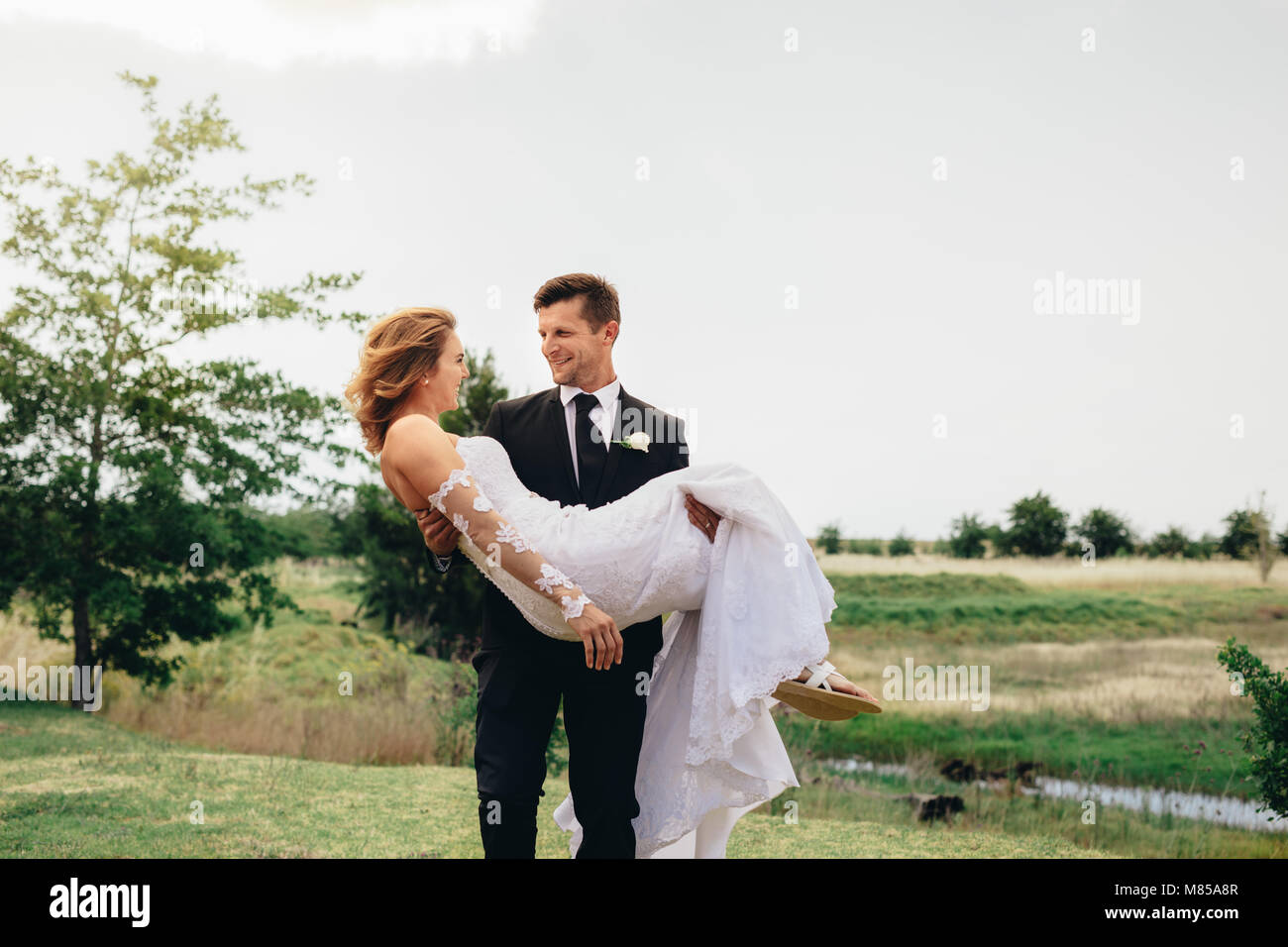 Young groom carrying his beautiful wife at the park. Bridegroom lifting ...