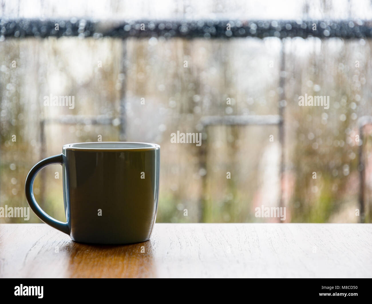 A mug on a wooden table in front of a glass window covered with rain ...