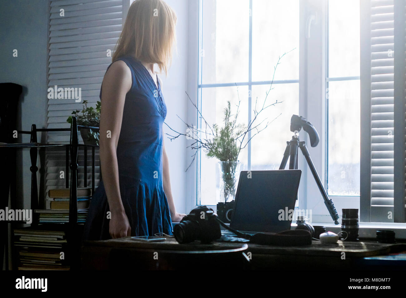 Mid adult woman, standing beside window, camera and laptop on table ...