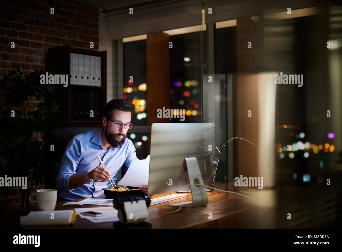 Young businessman reading paperwork and eating takeaway at office desk ...