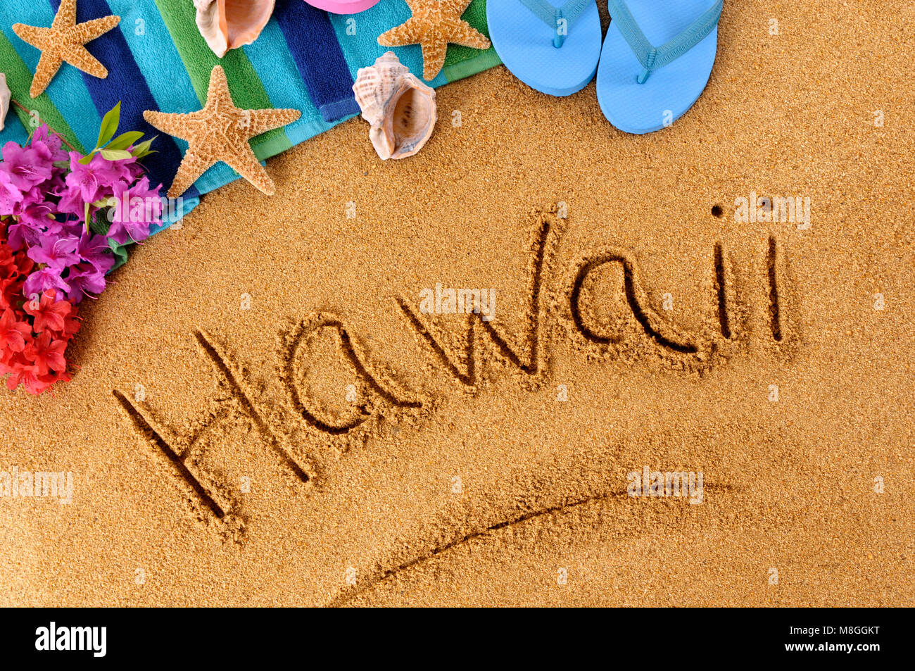 The word Hawaii written on a sandy beach, with flowers, beach towel ...