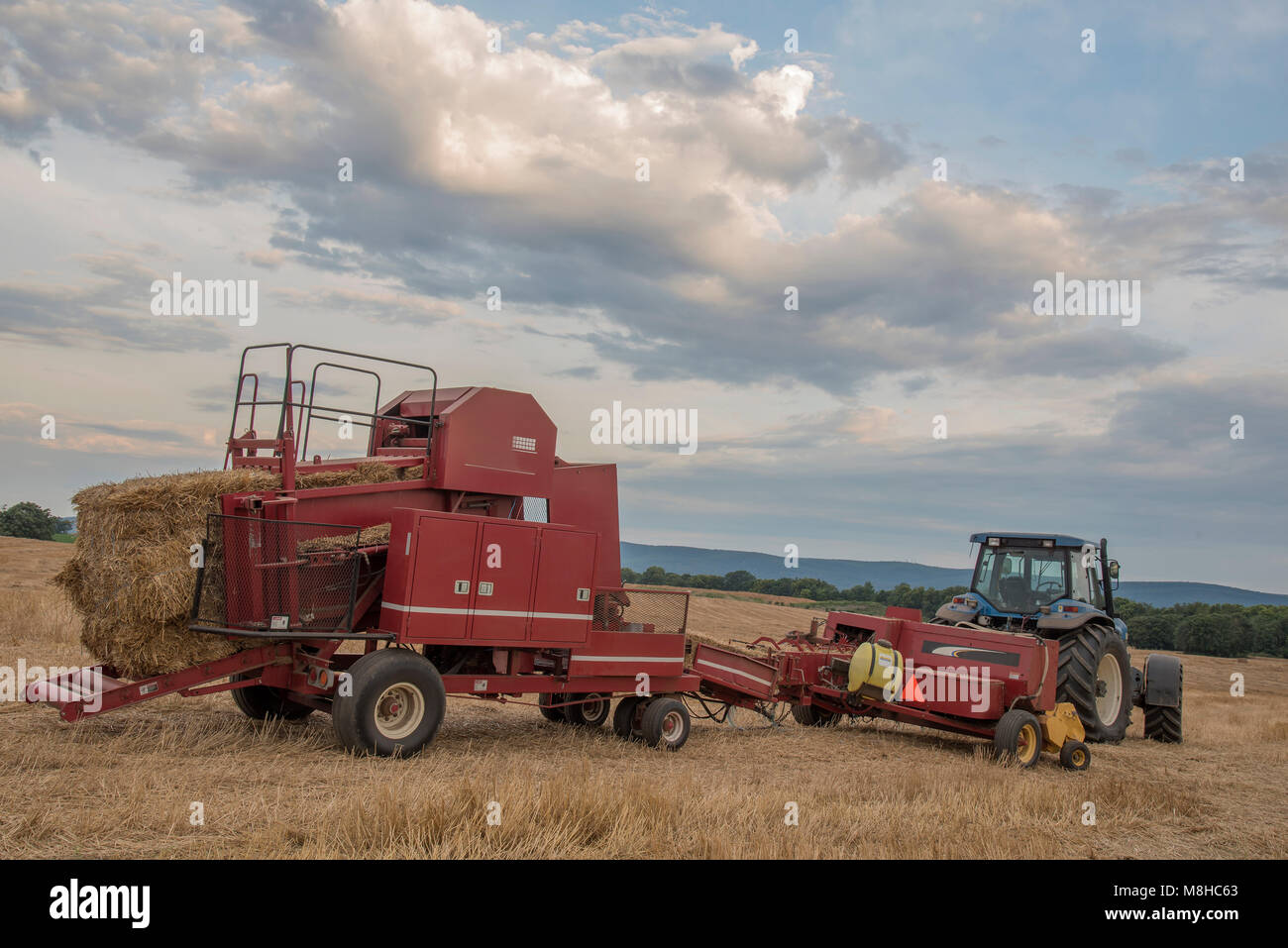 A blue tractor pulling a red hay bailer is parked for the night in a ...