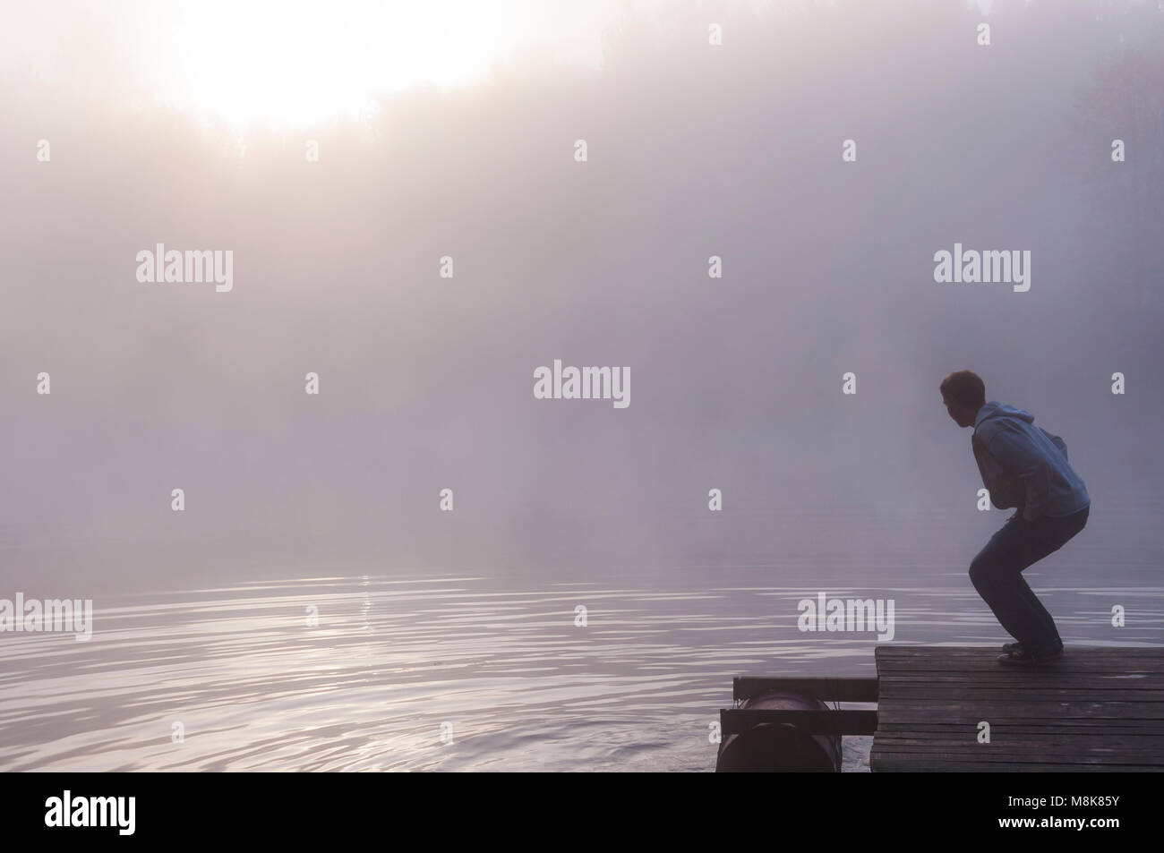 Young man making water ripples at foggy sunrise by jumping on lake pier ...