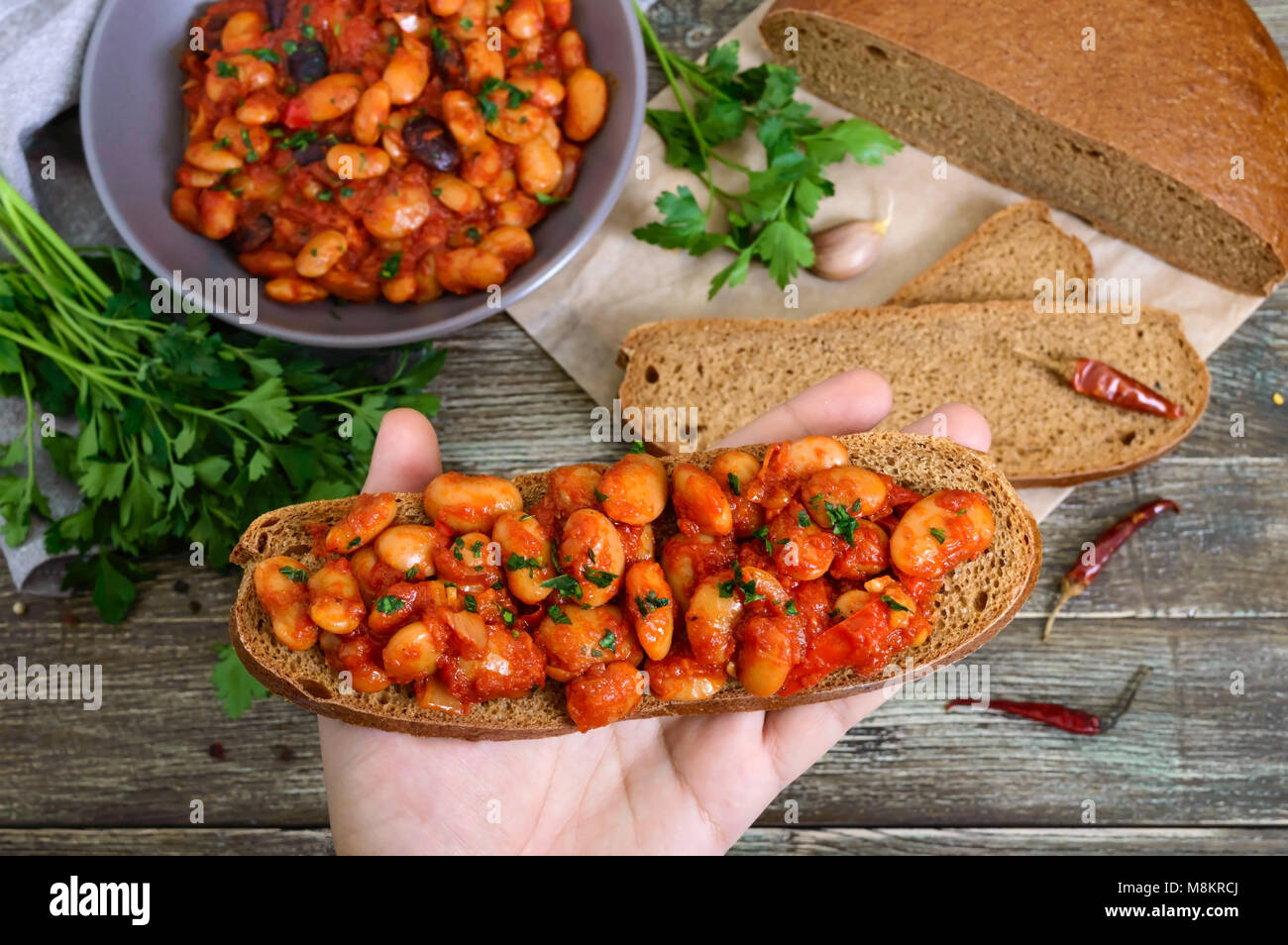 The hand holds a piece of rye bread with beans in the background of the ...