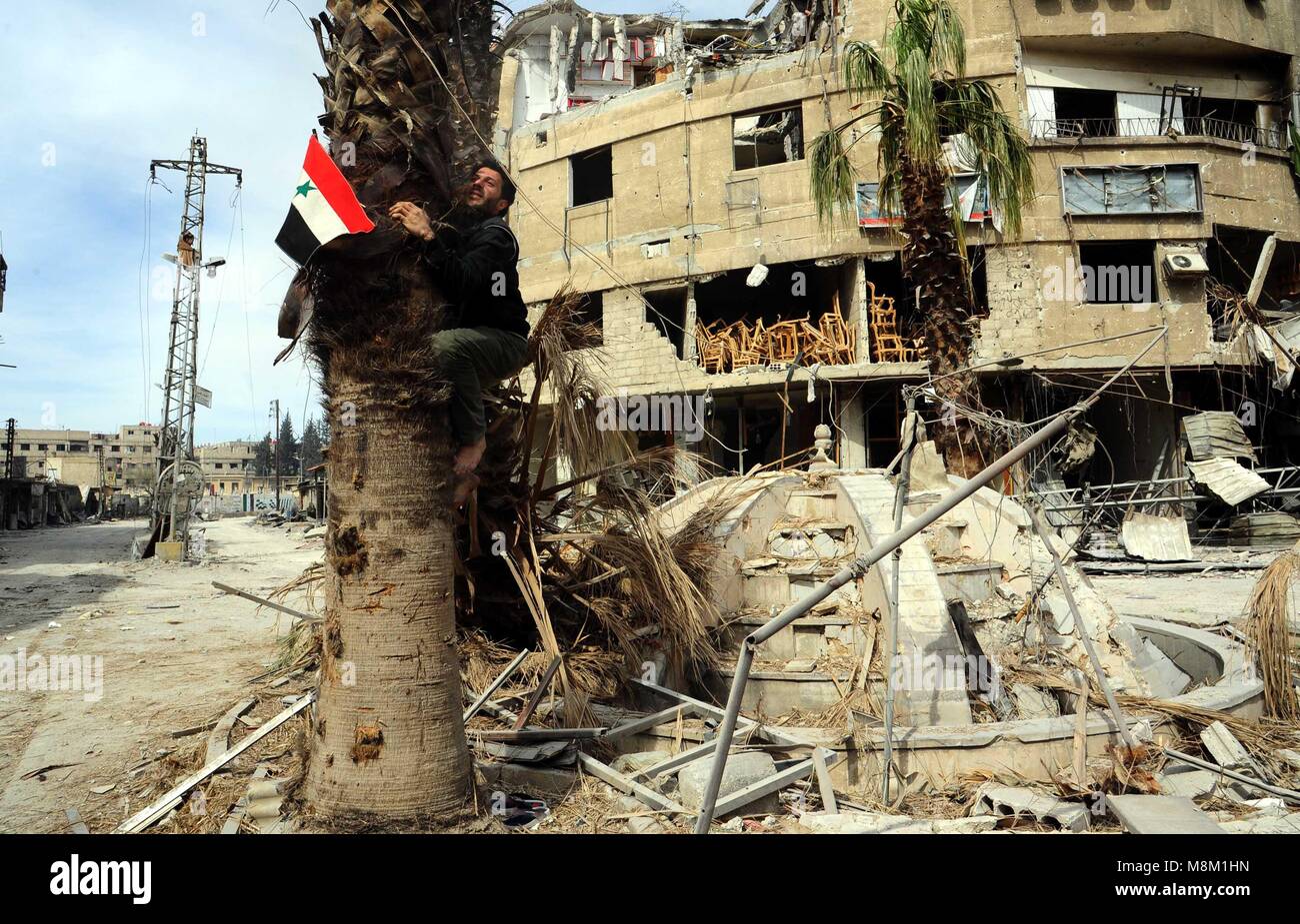 Damascus. 18th Mar, 2018. A man climbs a tree to hoist the Syrian flag ...