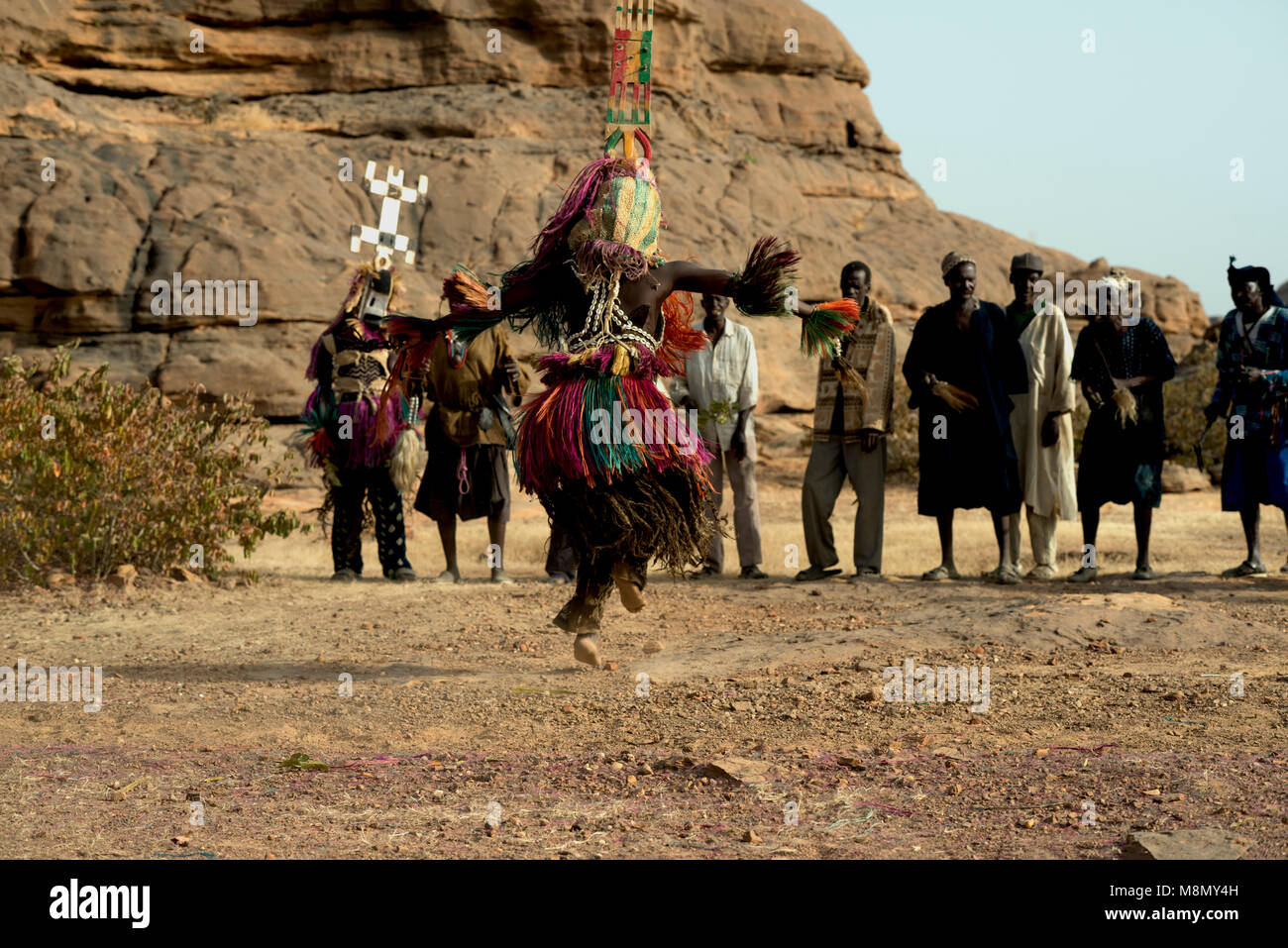 A group of Dogon masked dancers watch a man wearing a towering ...