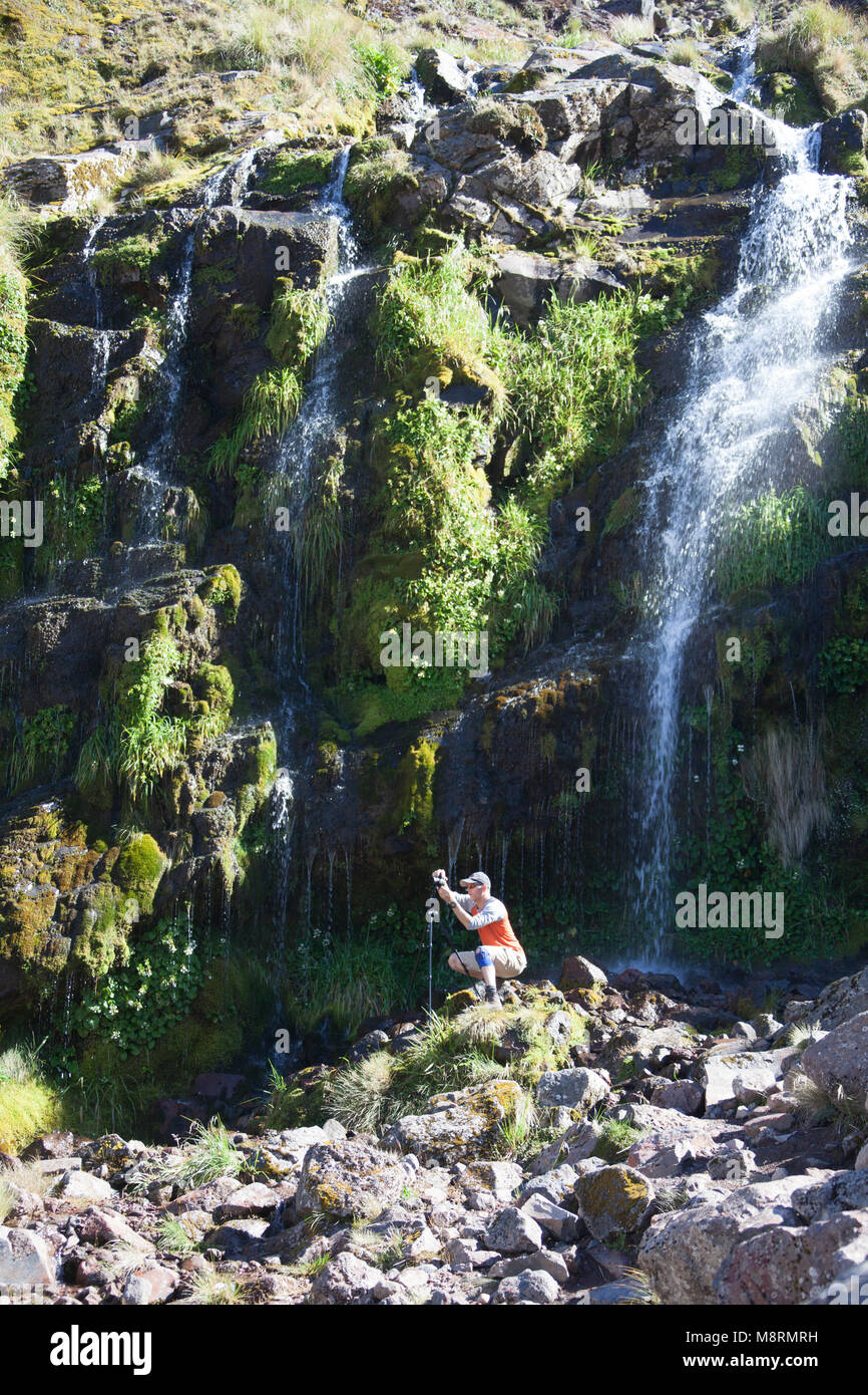 Hiker crouching on rocks against waterfall and mountain during sunny ...