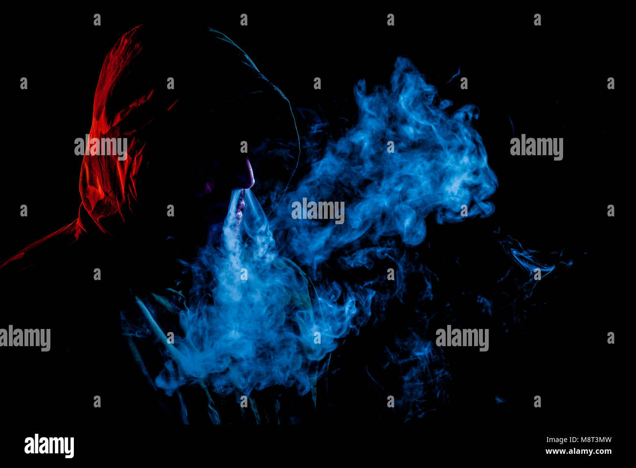 A young man in a black hood smokes a vape and exhales a colored red and ...