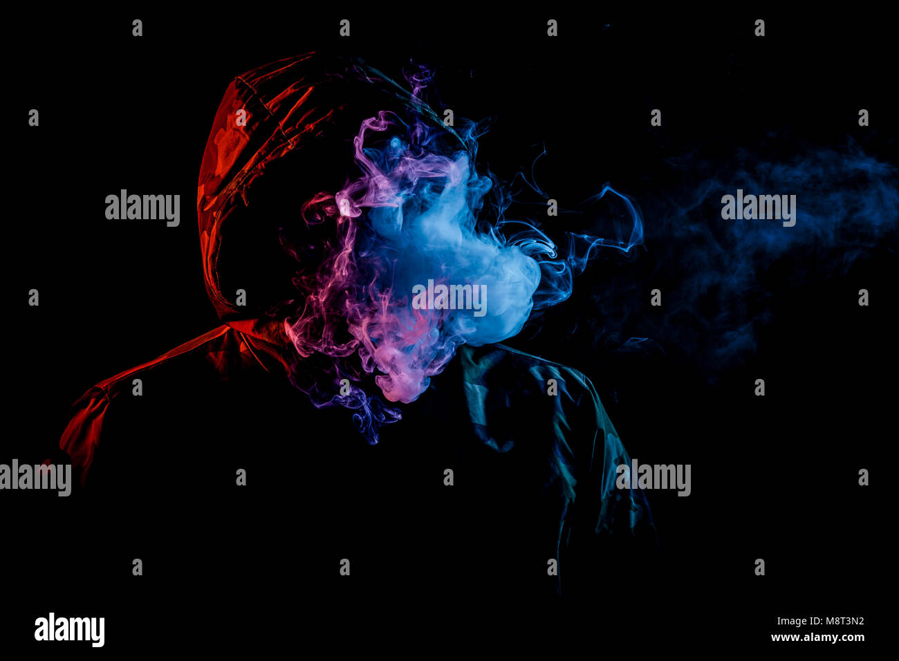 A young man in a black hood smokes a vape and exhales a colored red and ...