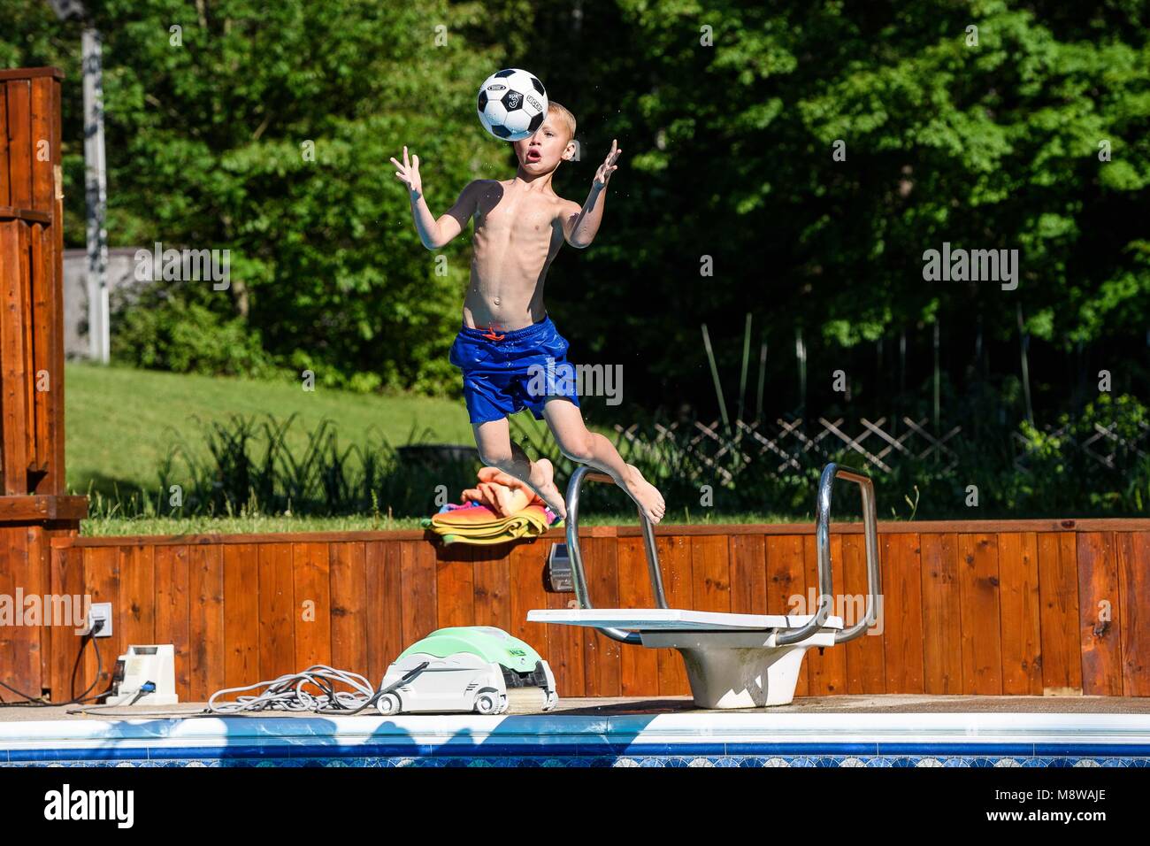 Blond boy catching soccer ball mid air while jumping into the pool ...