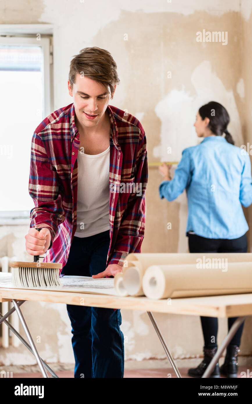 Young man applying adhesive to wallpaper while girlfriend is measuring ...
