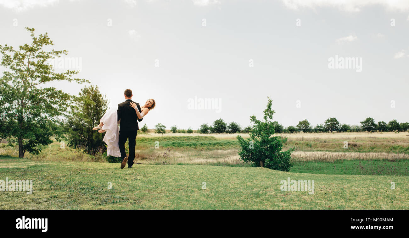Beautiful wedding couple with groom lifting up his bride at park. Rear ...