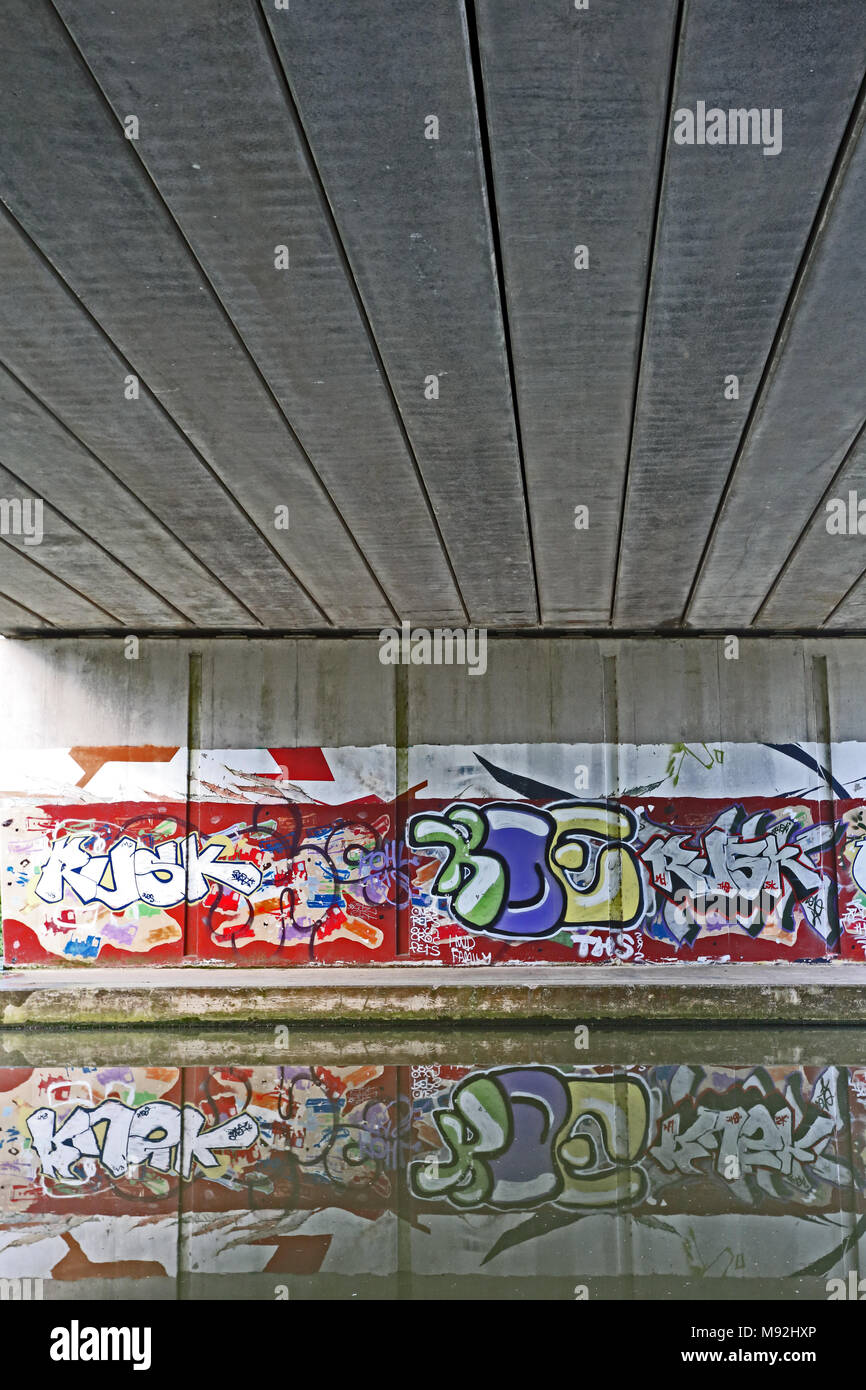 Graffiti beneath a road bridge is reflected in the calm water of the ...