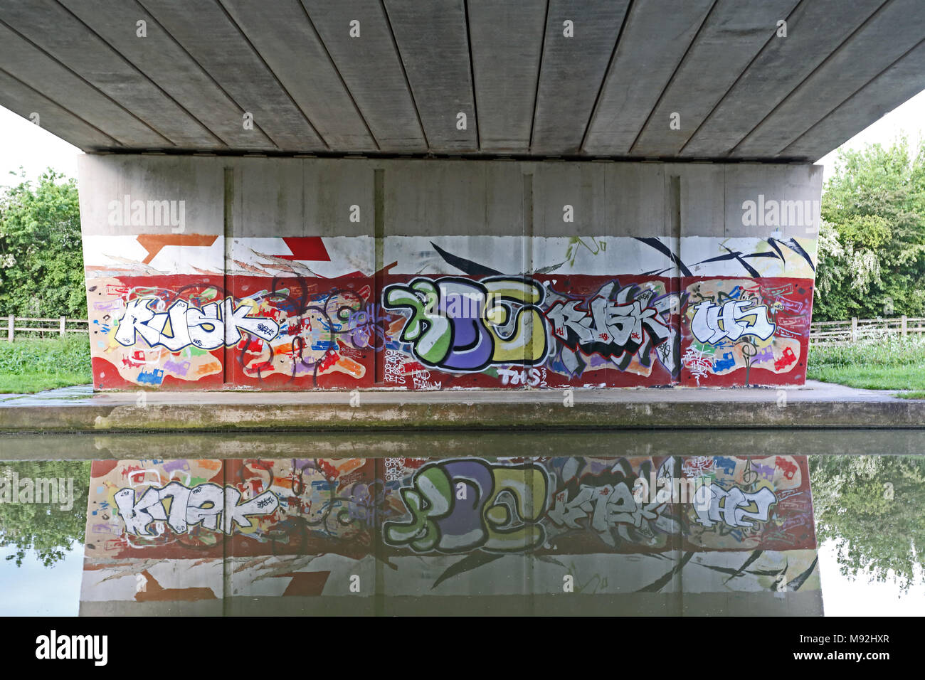 Graffiti beneath a road bridge is reflected in the calm water of the ...