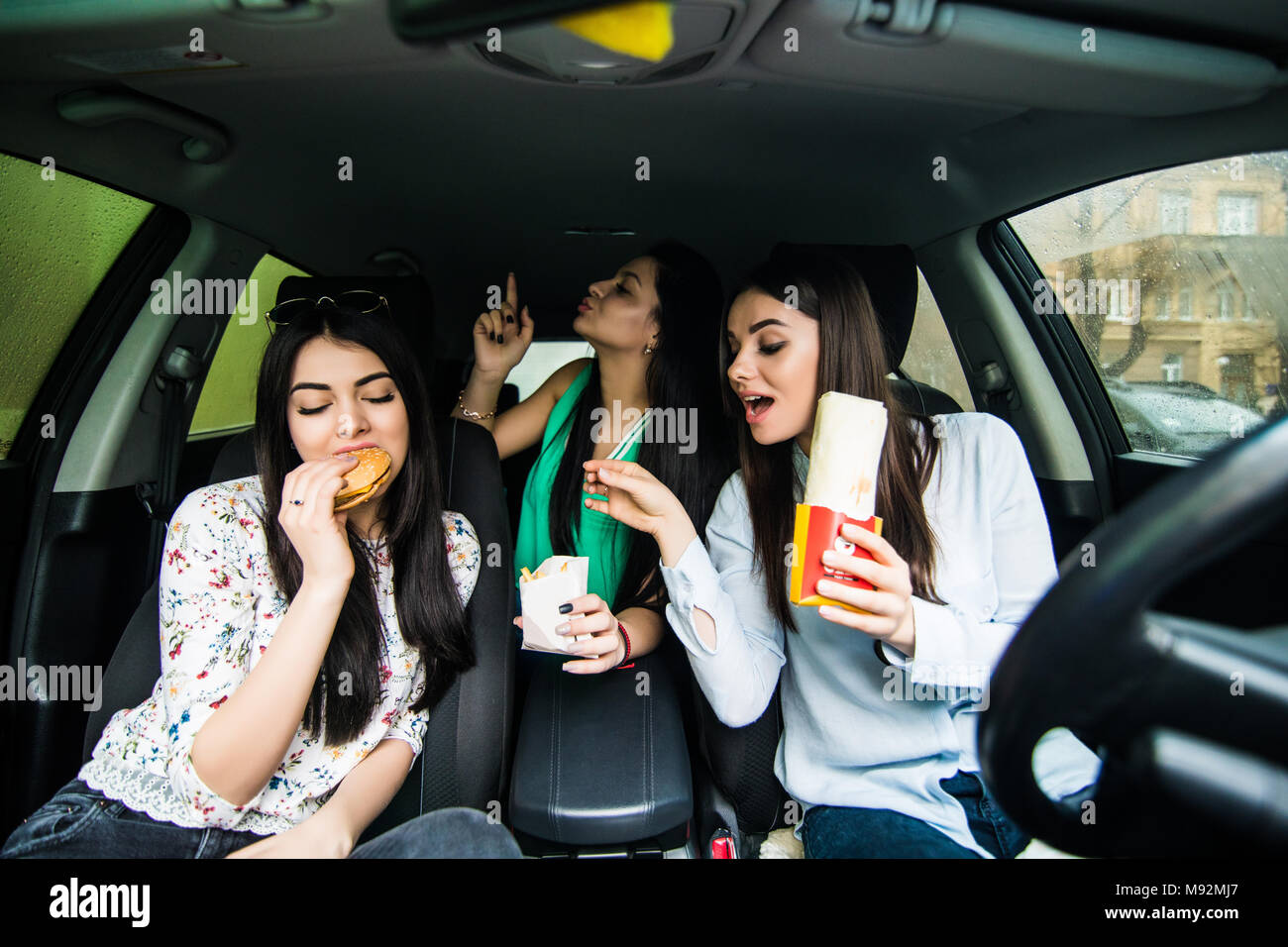 Three young women drive a car, speaking each other and eating a fast ...
